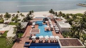 Área de piscina em resort com várias piscinas, espreguiçadeiras e guarda-sóis, cercada por palmeiras e vista para a praia.