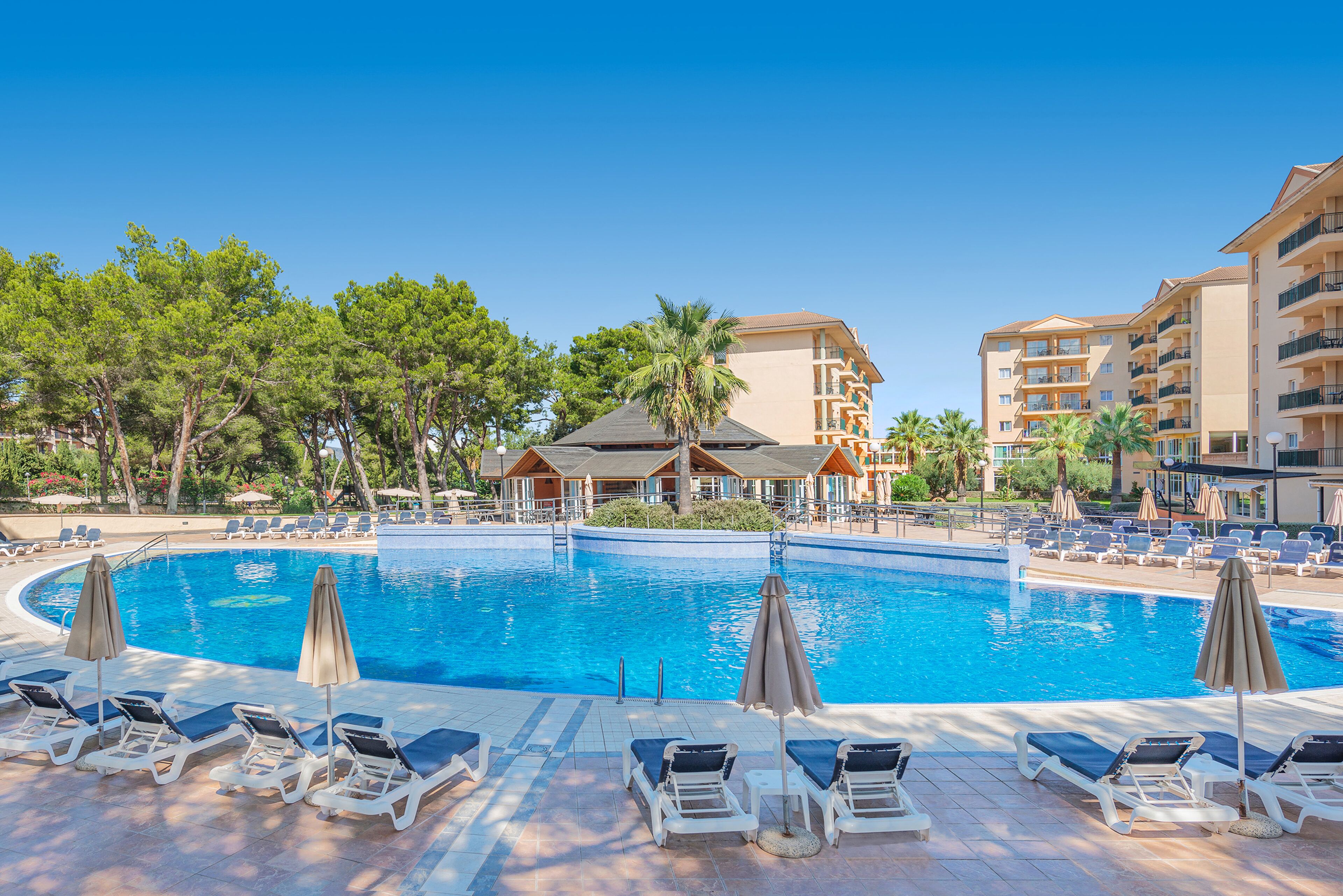 Piscina com espreguiçadeiras e guarda-sóis, cercada por árvores e edifícios de hotel.