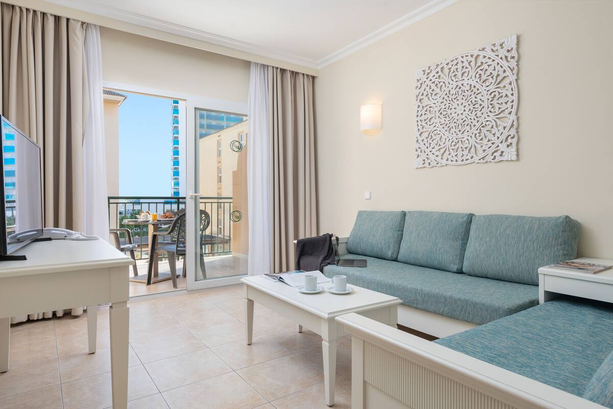 Quarto de hotel moderno com varanda, sofá, mesa de jantar e um detalhe decorativo na parede.