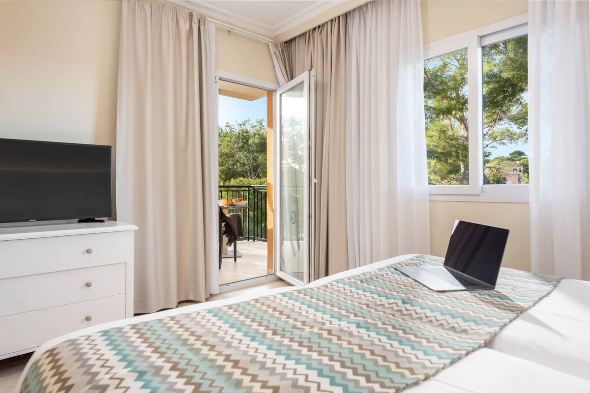Quarto de hotel com cama, televisão, laptop, janela com vista para árvores e varanda.
