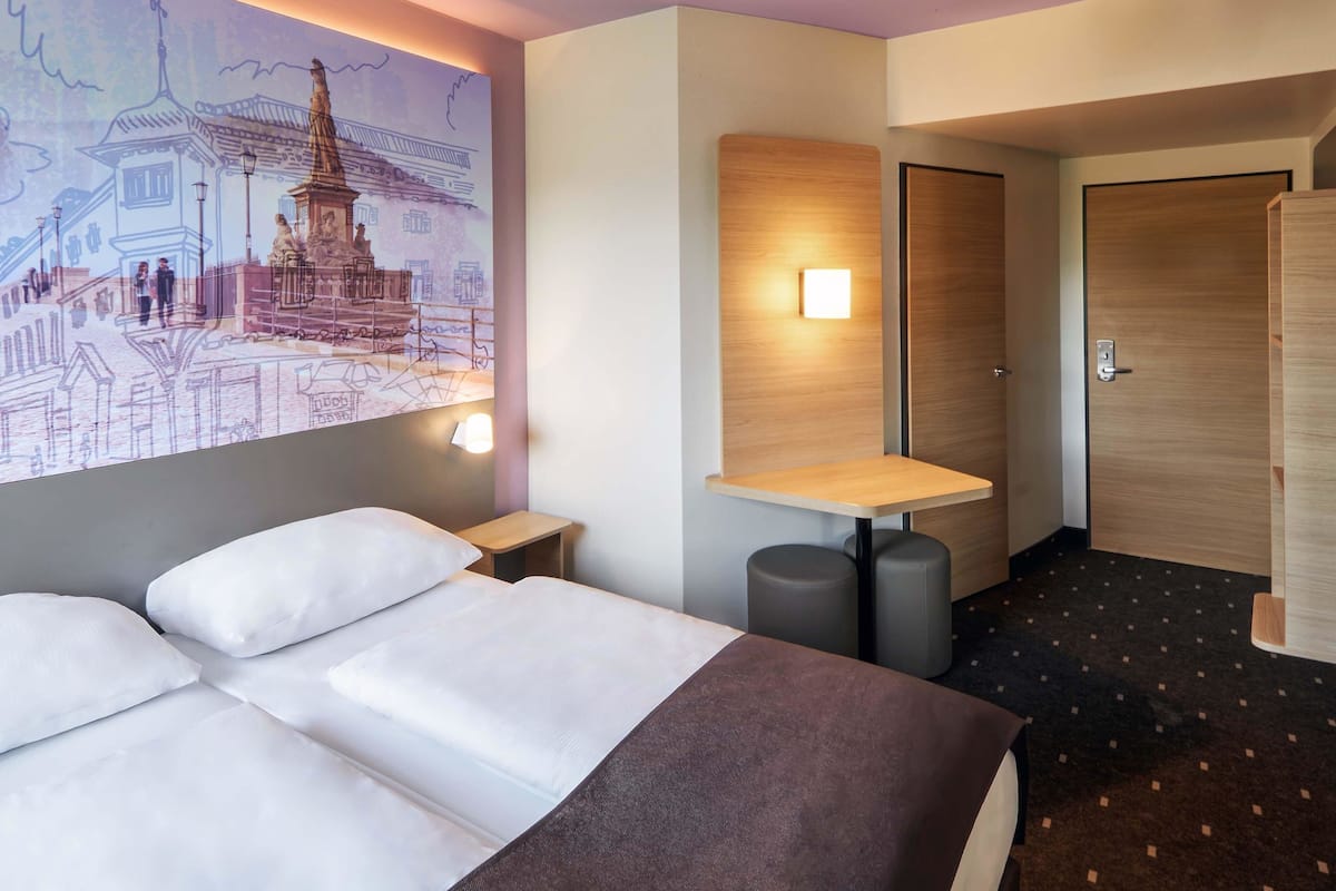 Quarto de hotel com uma cama, uma mesinha, uma luminária na parede e uma porta.
