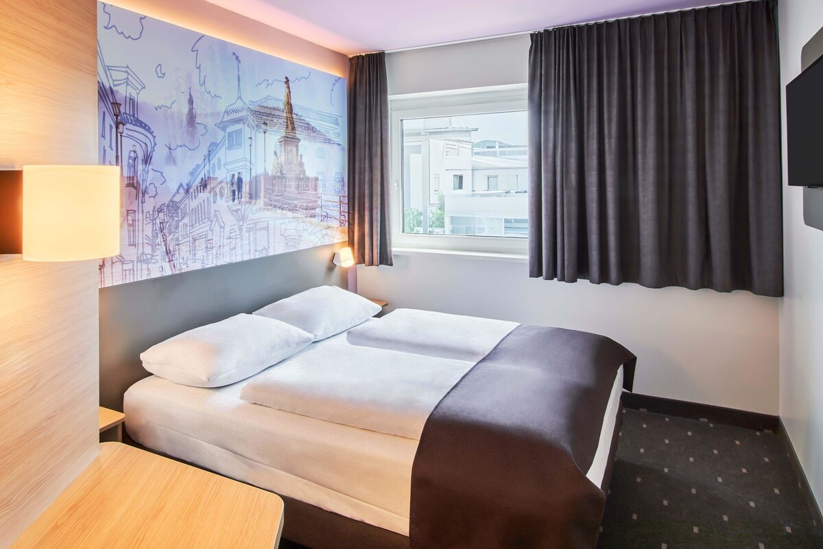 Quarto de hotel com cama, escrivaninha, luminária, pintura de uma paisagem urbana, janela com cortinas e televisão na parede.