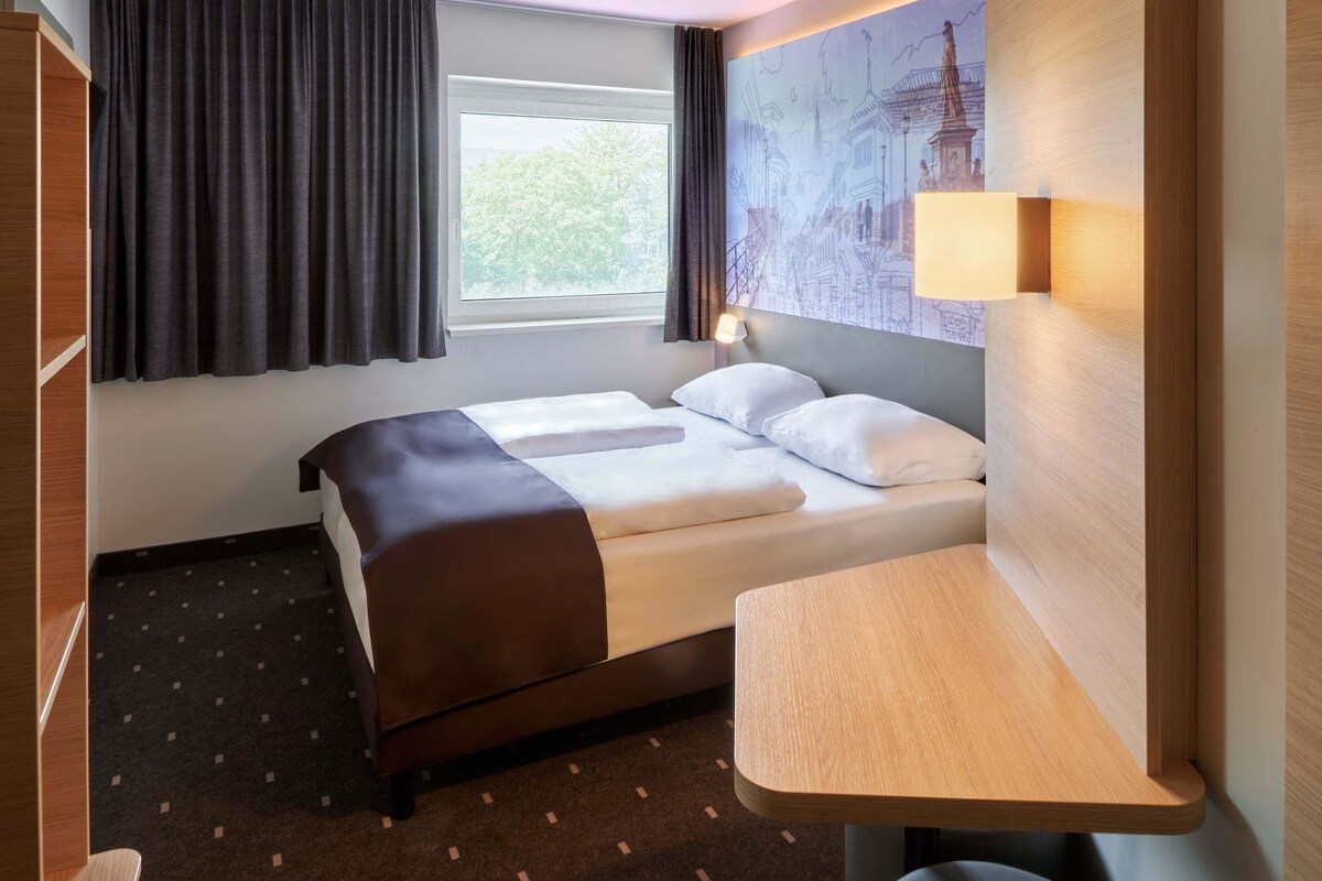 Quarto de hotel com uma cama, uma escrivaninha, janela com cortinas e parede com um mural da paisagem urbana.