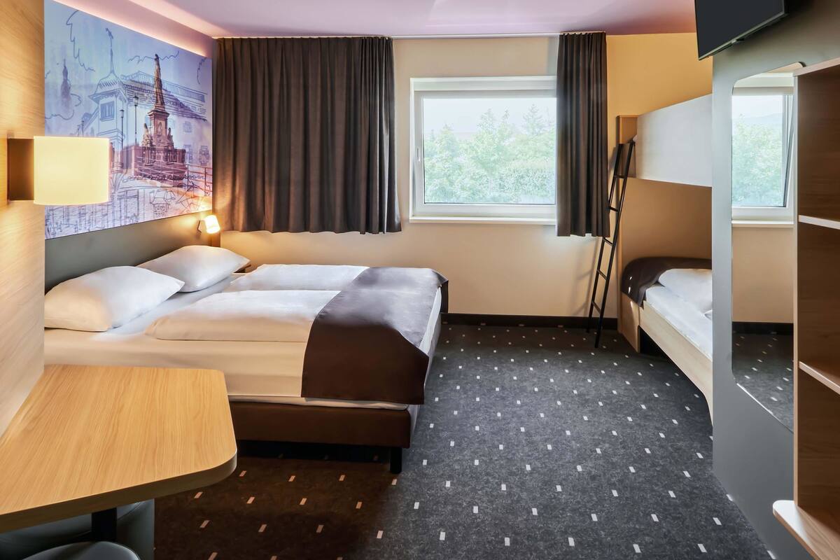 Quarto de hotel com cama, escrivaninha, cadeira, televisão e janela com cortinas.