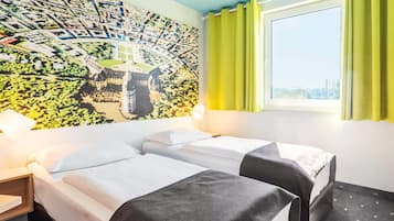 Quarto de hotel com duas camas, uma janela grande com cortinas, um mural com vista para a cidade na parede e uma mesa de cabeceira com abajur.