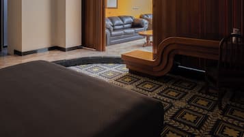 Quarto de hotel com uma cama grande, um banco de madeira, uma mesa pequena e um sofá ao fundo.