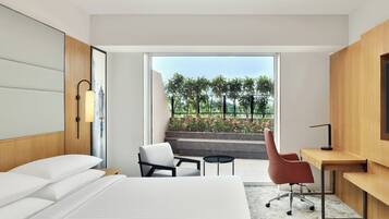 Quarto de hotel moderno com uma cama grande, uma escrivaninha, uma cadeira e vista para um jardim e árvores através de uma janela ampla.