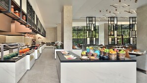 Um hall de hotel moderno com uma ilha central exibindo frutas, uma área de cozinha com buffet e um espaço de estar com mesas e cadeiras.