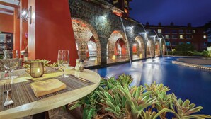 Uma área de refeições à beira da piscina com parede de pedra e aberturas em arco, mesa posta com um jogo de mesa e piscina com cascata.