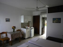 Quarto compacto com cama, pequeno espaço de cozinha, mesa de jantar com cadeiras e ventilador de teto.