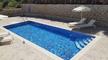 Uma piscina retangular com água azul cristalina, rodeada por um muro de pedra e piso de azulejo. Duas cadeiras de descanso brancas e um guarda-sol branco estão próximos à piscina.