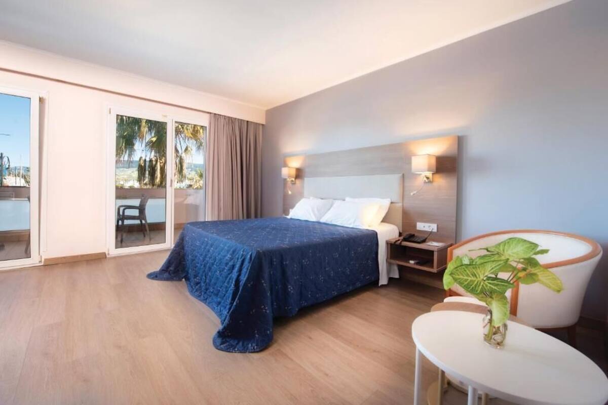 Quarto de hotel com cama, mesa de cabeceira, luminária e vista para palmeiras pela janela.