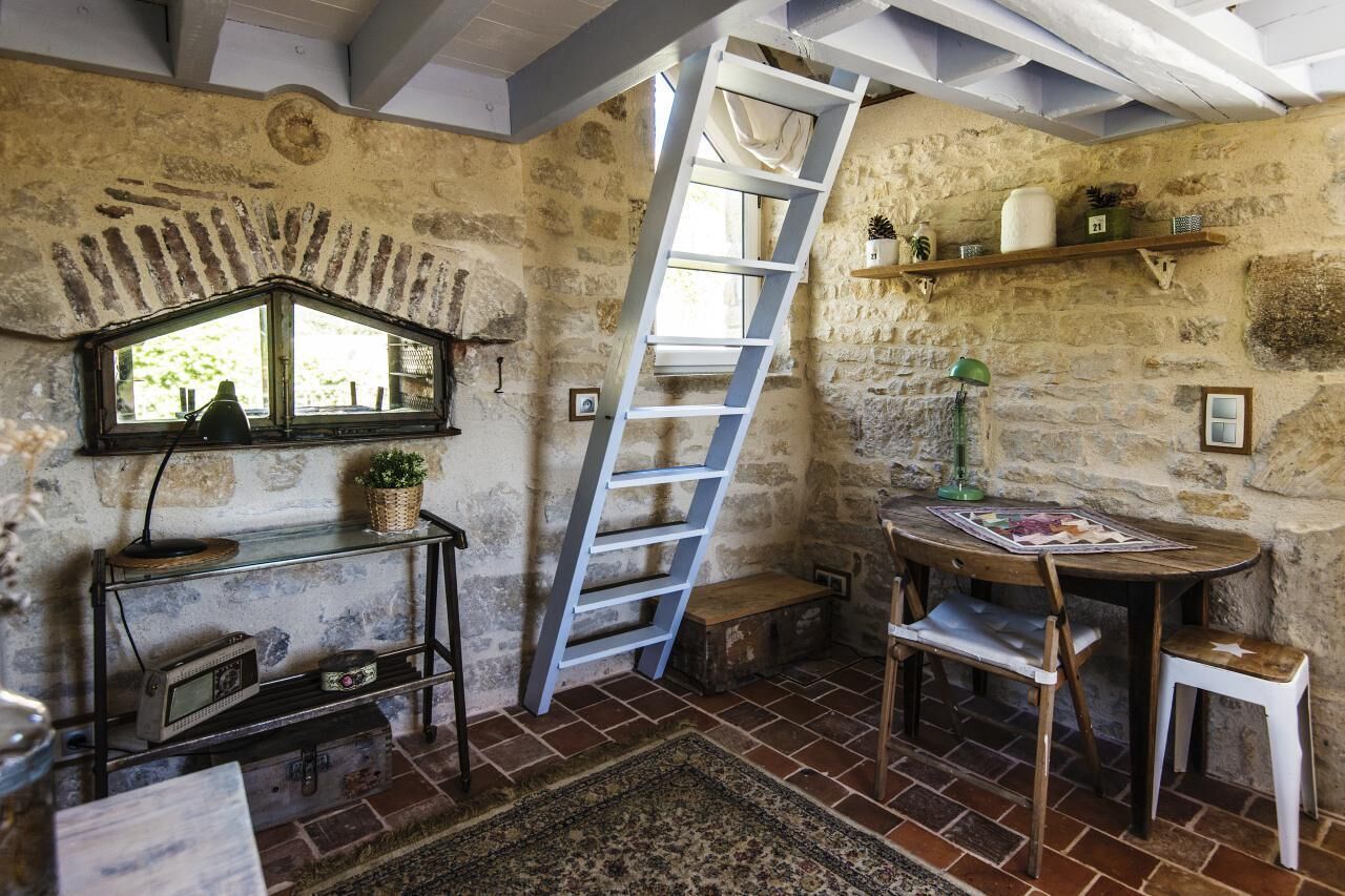 Quarto rústico com parede de pedra, mesa de madeira, escada e prateleira com objetos decorativos.