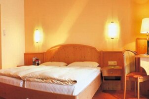 Quarto de hotel com cama de madeira, duas luminárias na parede, uma mesa de cabeceira e uma cadeira.