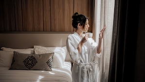Uma pessoa vestida com um robe branco, em pé perto de uma janela com cortinas, uma cama com roupa de cama branca e almofadas estampadas, e painéis de madeira na parede.