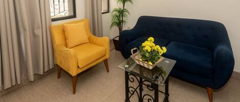 Uma sala de estar com uma poltrona amarela, um sofá azul e uma mesa de centro de vidro com uma planta.