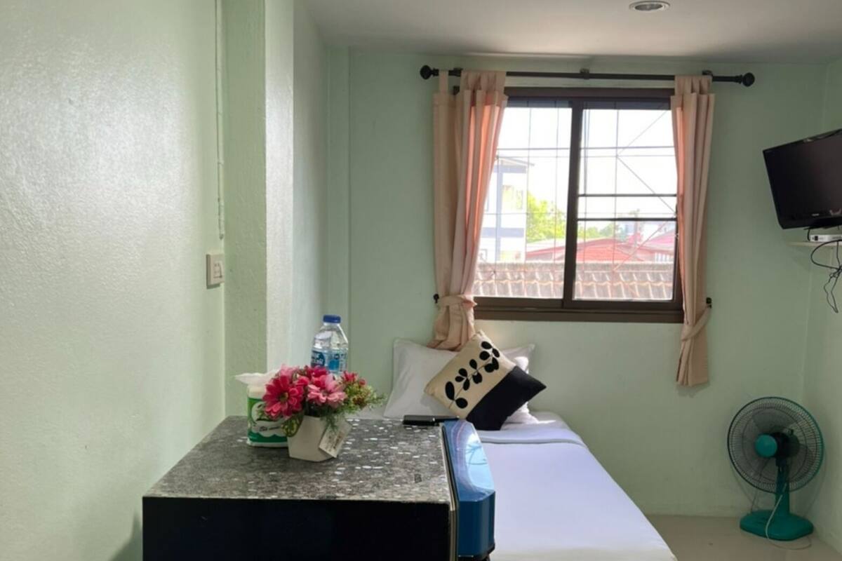Quarto pequeno e limpo com uma cama, escrivaninha com cadeira, janela com cortinas, televisão na parede e ventilador.