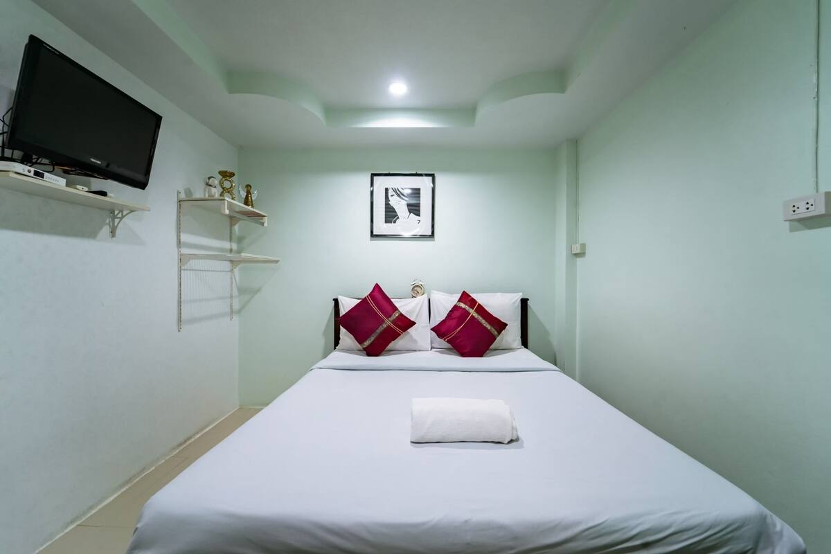 Uma cama bem arrumada com almofadas brancas e vermelhas, uma TV de tela plana na parede e um quadro emoldurado sobre a cama.