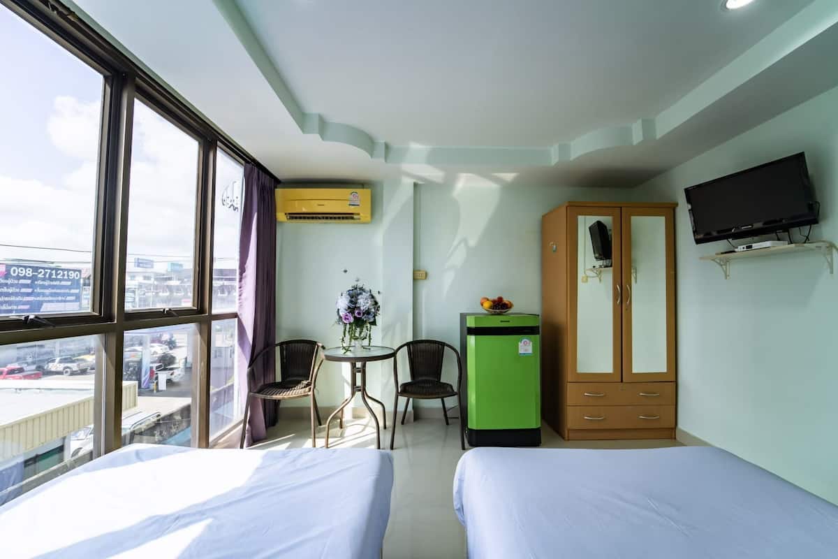 Quarto de hotel com duas camas, um minibar verde, televisão, guarda-roupa de madeira, mesa com cadeiras e janela com vista para a cidade.