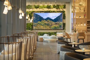 Interior moderno com uma TV grande fixada na parede exibindo uma paisagem montanhosa, áreas de estar com poltronas estofadas e mesas, e um bar com prateleiras cheias de garrafas e copos.