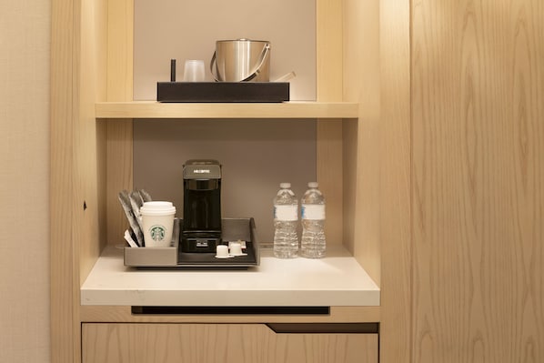 Minibar de hotel com cafeteira, copo da Starbucks, garrafas de água e bandeja com utensílios.