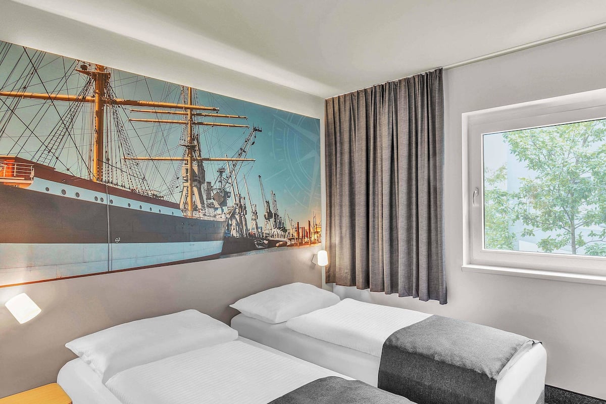 Quarto de hotel com duas camas, uma pintura grande de um navio na parede, uma janela com cortinas e uma mesinha de cabeceira.