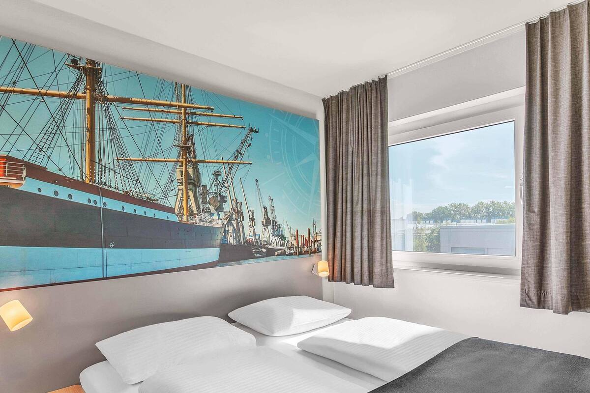 Quarto de hotel com uma cama grande, uma mesa de cabeceira, uma janela com cortinas e um mural na parede com o tema de um navio.