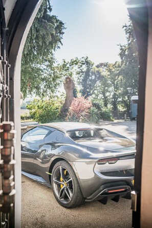 Um carro esportivo estacionado com um spoiler traseiro visível e pinças de freio amarelas, em uma entrada de garagem com uma árvore e um poste de luz ao fundo.