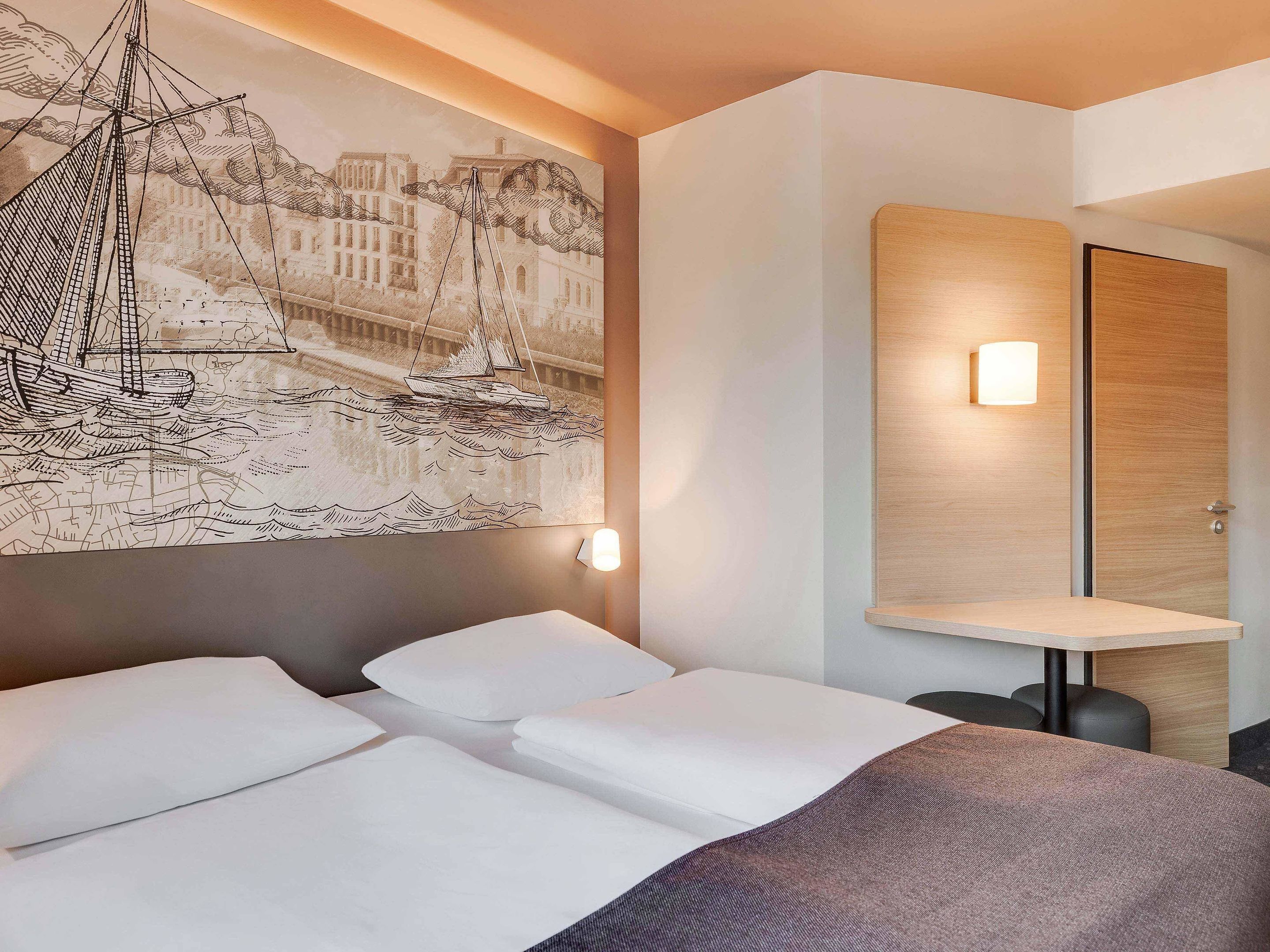 Quarto de hotel com uma cama, uma obra de arte pendurada na parede retratando um veleiro, uma porta de madeira e uma mesinha de cabeceira com abajur.