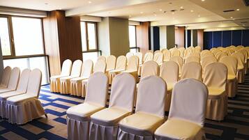Uma sala de conferência com fileiras de cadeiras e mesas, janelas amplas e um projetor no teto.