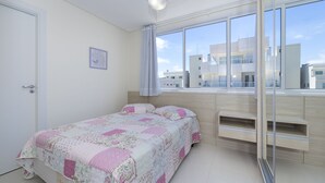 Quarto com cama, janela com vista para edifícios, porta e ventilador de teto.