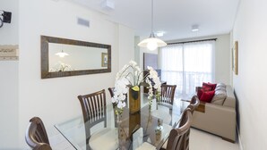 Uma área de jantar com mesa de vidro, cadeiras e um vaso de flores. Um sofá com almofadas vermelhas ao fundo.