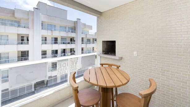 Varanda com mesa e cadeiras de madeira, televisão na parede e vista para um edifício alto.