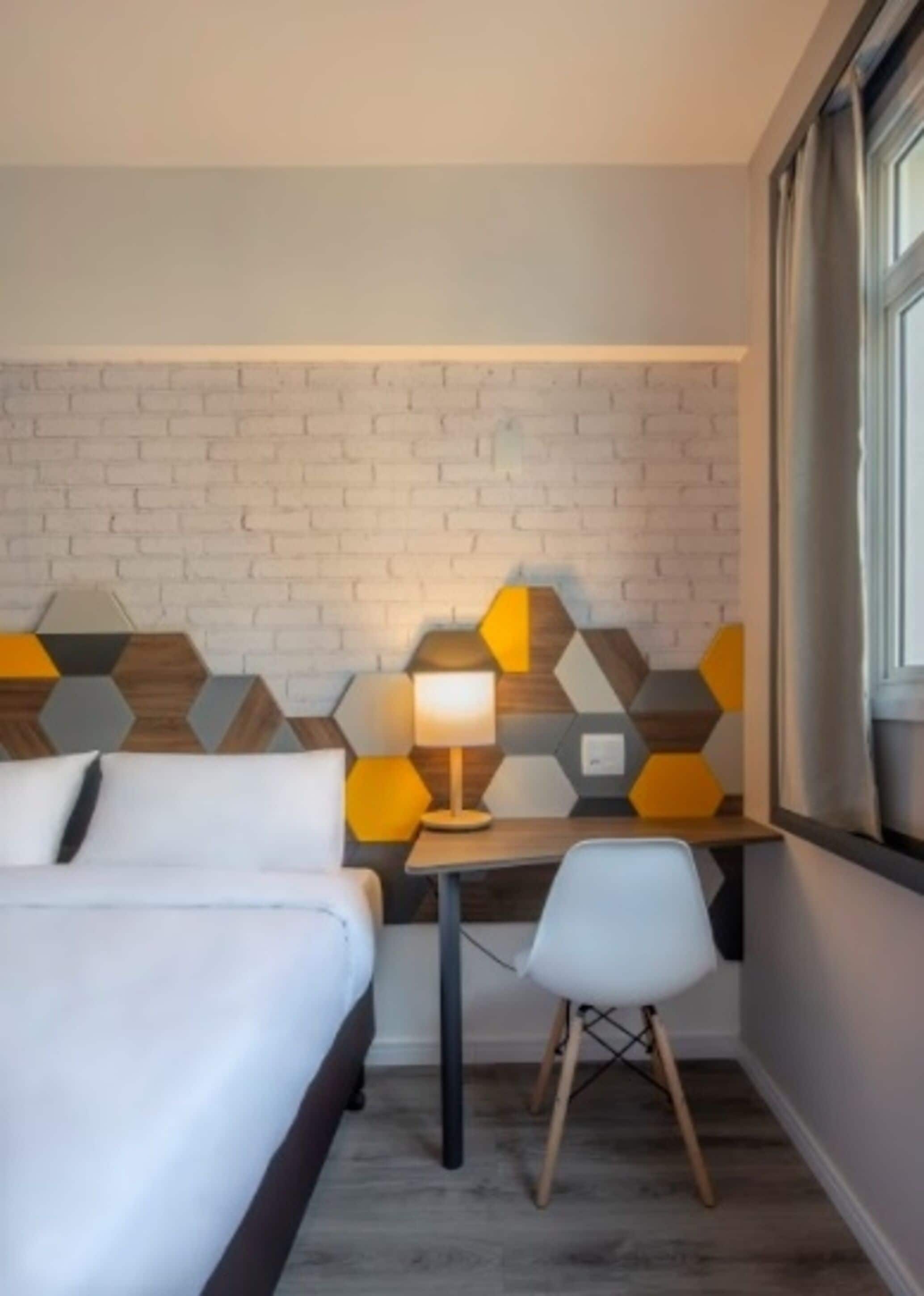 Quarto de hotel com parede em padrão de tijolos, uma escrivaninha com abajur, uma cadeira e duas camas.