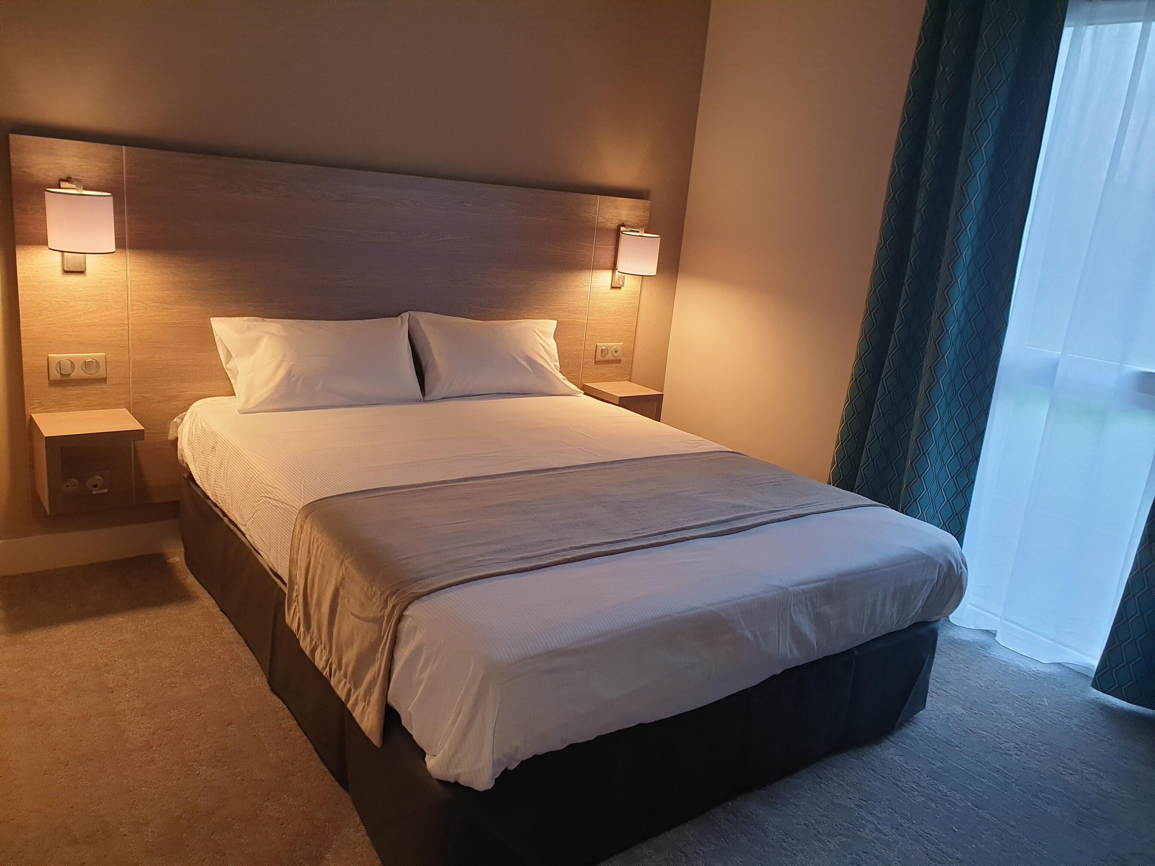Cama bem arrumada com lençóis brancos, duas mesas de cabeceira com abajures e vista para uma janela com cortinas.