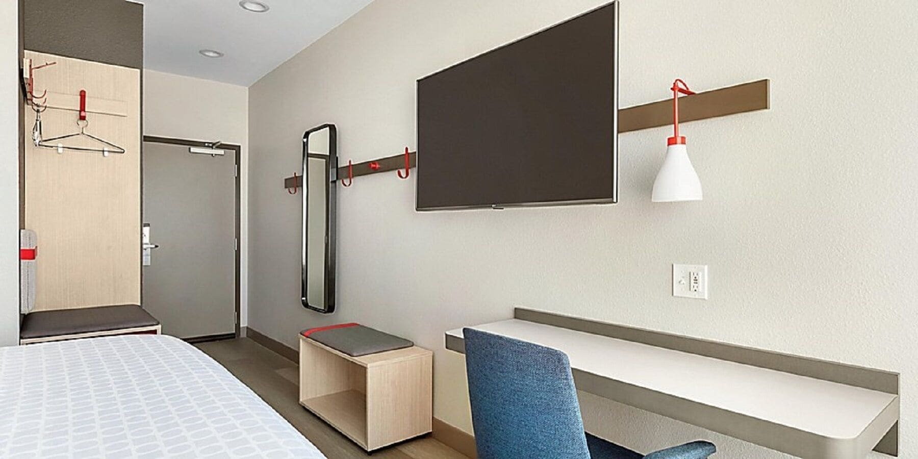Quarto de hotel com cama, escrivaninha, cadeira, televisão fixada na parede, espelho e luminária suspensa.