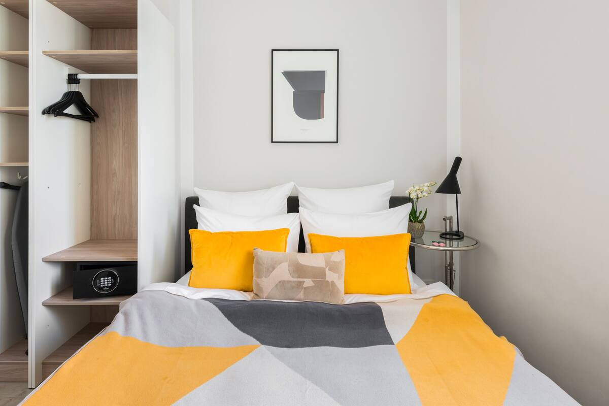 Quarto bem arrumado com uma cama, mesa de cabeceira com abajur, quadro na parede e guarda-roupa com roupas penduradas.