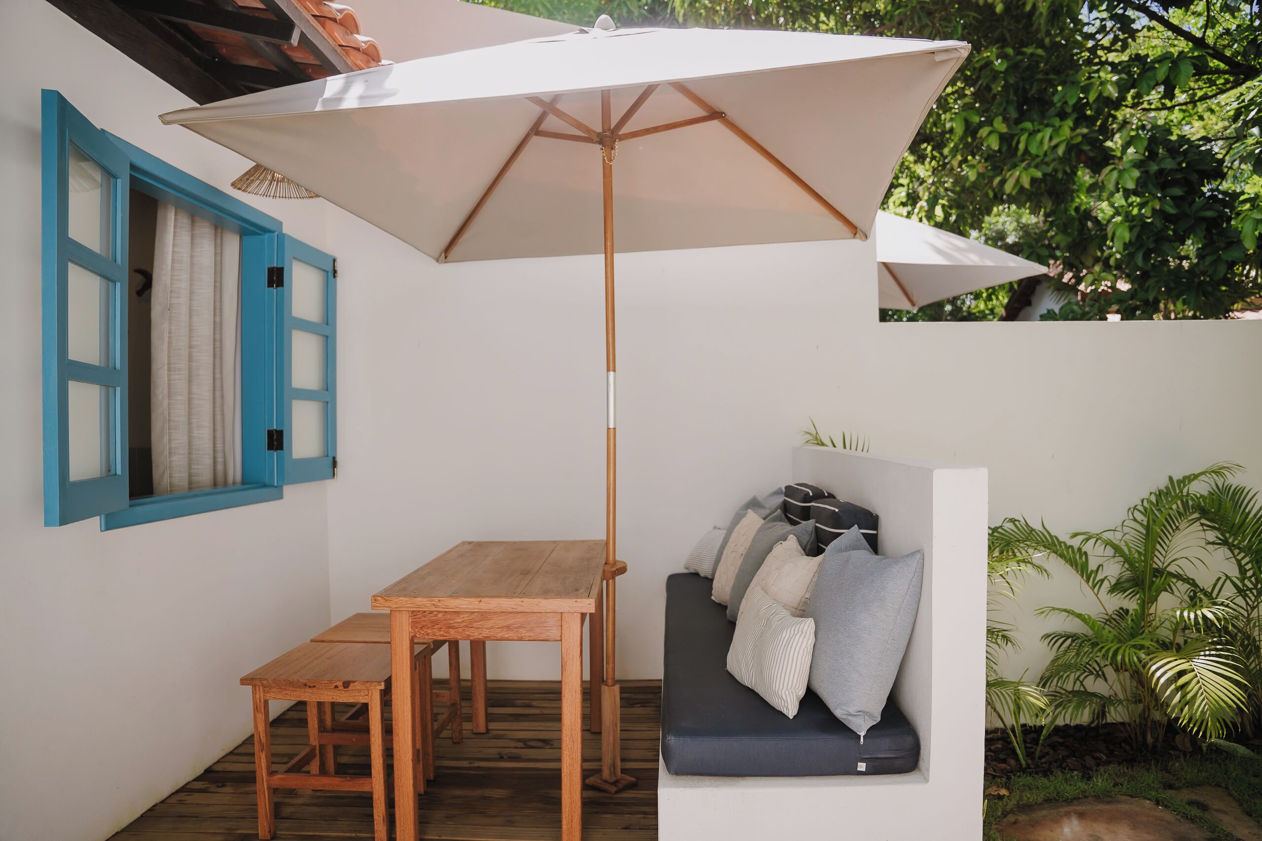 Uma mesa e banco de madeira sob um guarda-sol branco, uma janela com moldura azul e um sofá com almofadas.