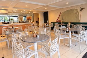 Uma área de refeições de hotel com mesas e cadeiras redondas, uma cozinha ao fundo e um mural na parede.