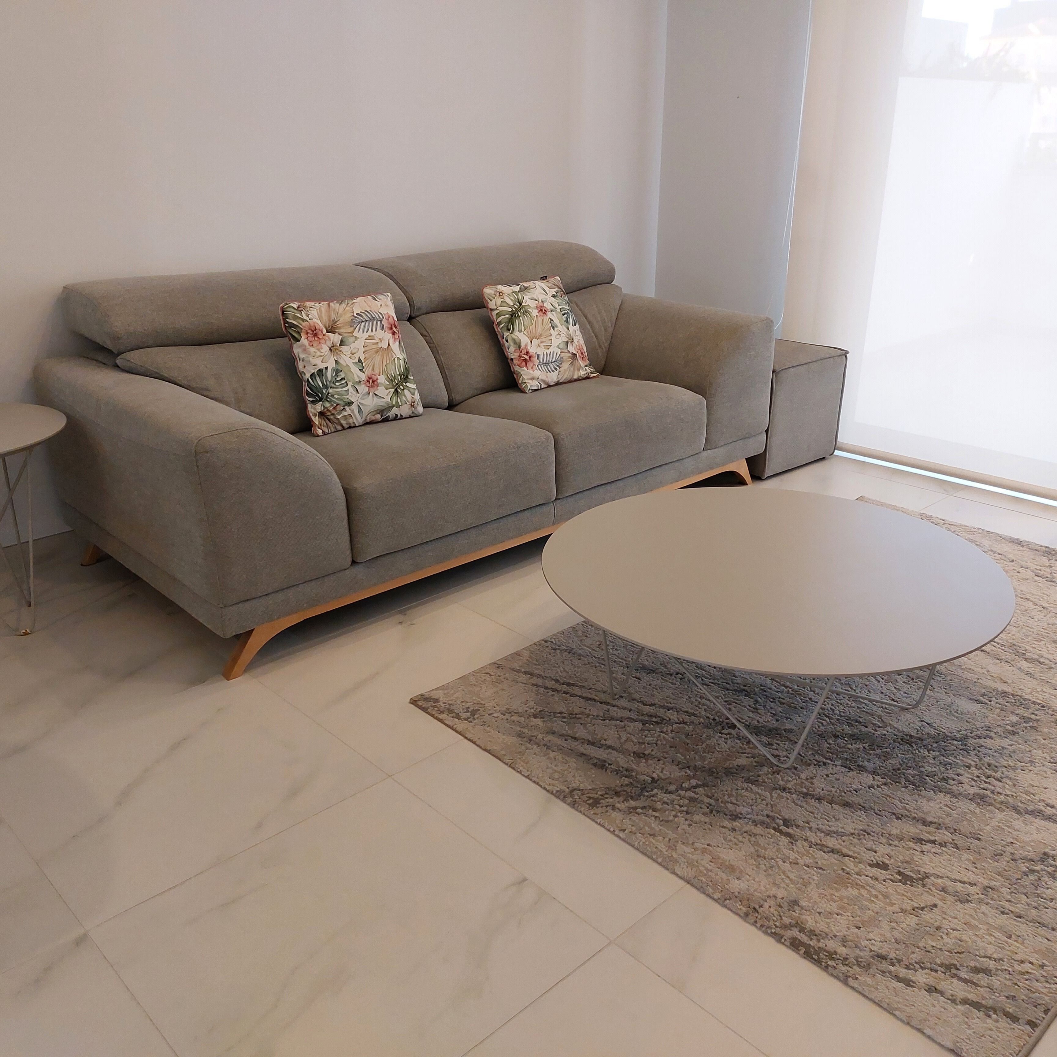Uma sala de estar moderna com um sofá cinza, duas almofadas florais, uma mesa de centro redonda e um tapete estampado.