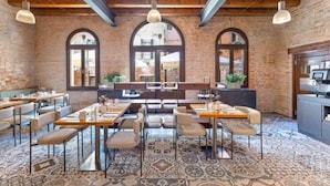 Restaurante com paredes de tijolo, janelas em arco e piso estampado. Mesas e cadeiras dispostas para refeições.