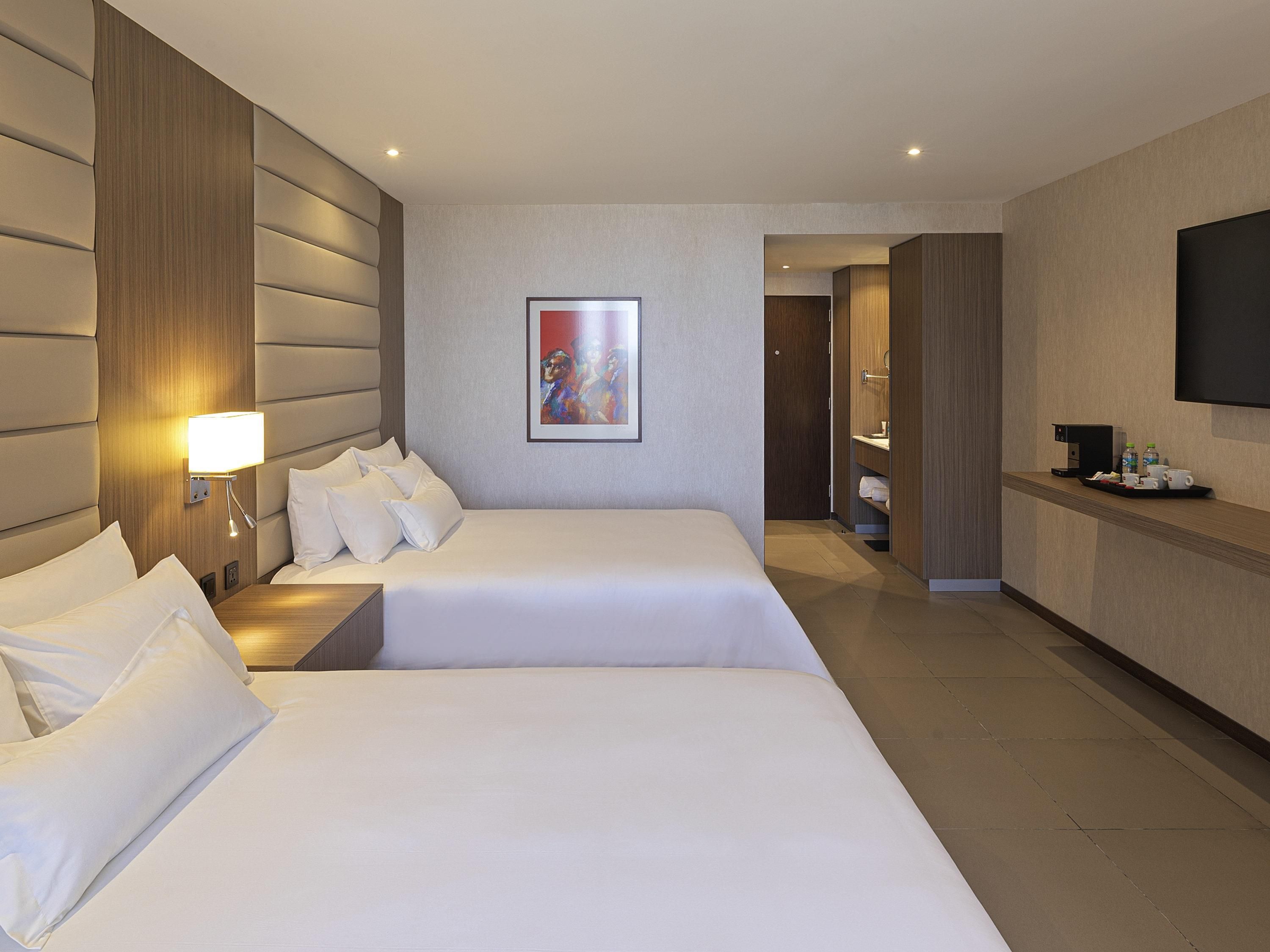 Quarto de hotel com duas camas, um quadro na parede, uma televisão e uma mesa com cafeteira.
