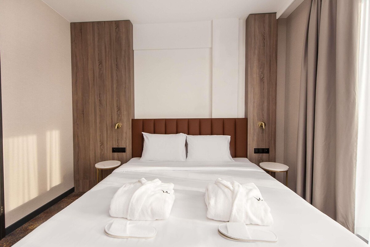Quarto de hotel com uma cama grande, duas mesas de cabeceira, um painel de cama em madeira e vista para o exterior.