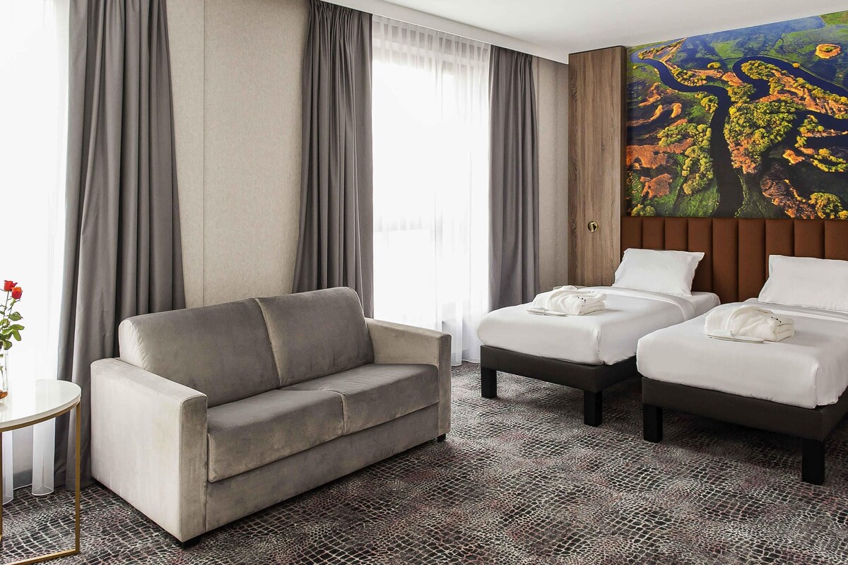 Quarto de hotel com sofá, duas camas, um quadro e um tapete estampado.