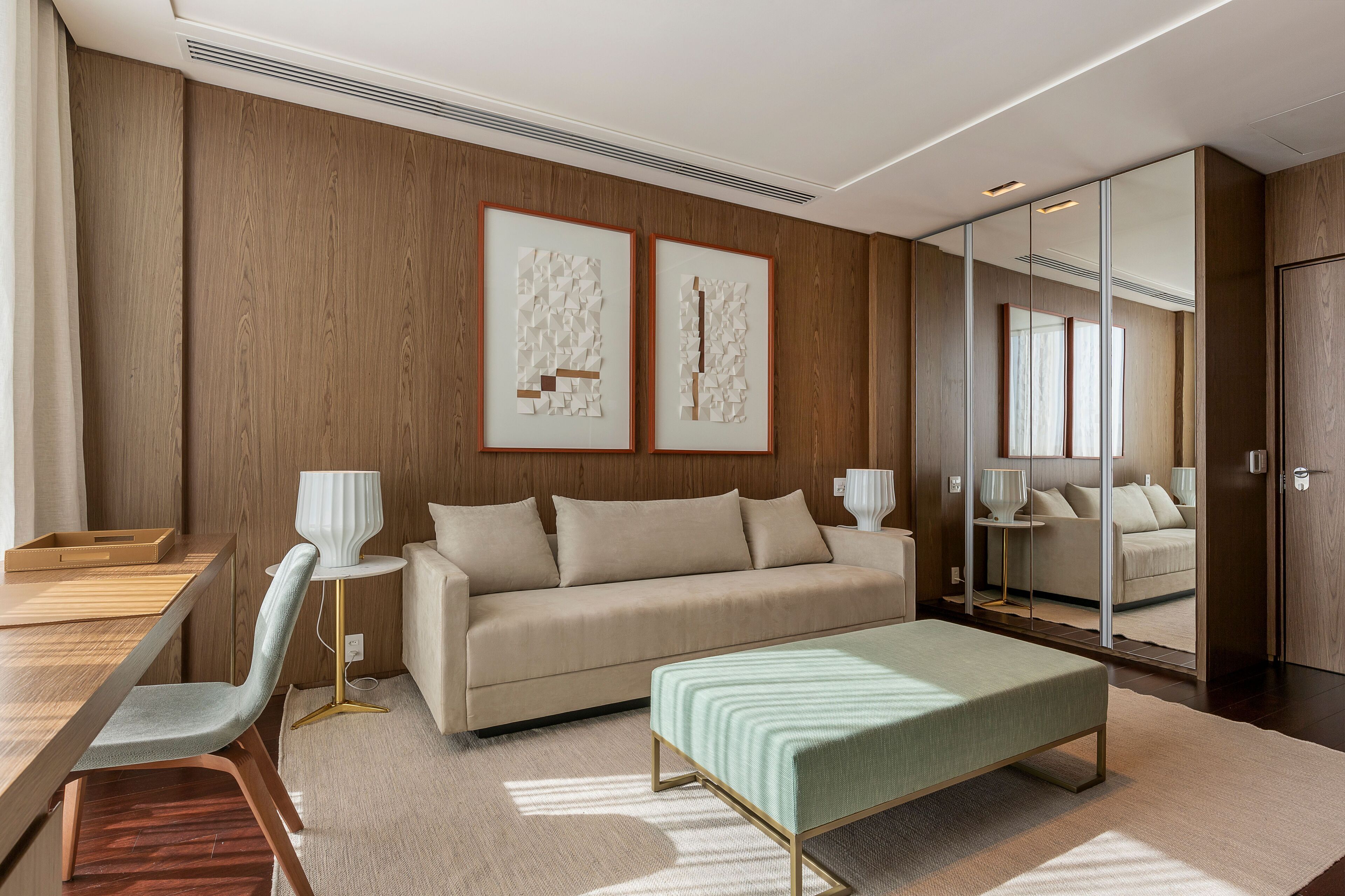 Quarto de hotel moderno com parede de painel de madeira, sofá, mesa de jantar com cadeiras, mesa de cabeceira com luminárias e um espelho grande.