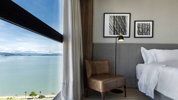 Quarto de hotel com uma janela grande, uma cama, uma cadeira e duas fotos emolduradas na parede.