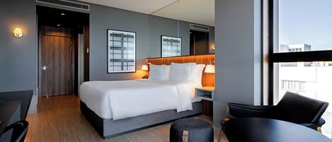 Quarto de hotel moderno com cama, mesa de cabeceira, cadeira, banquinho e uma janela grande.