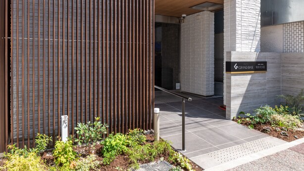Entrada de um edifício moderno com um portão de ripas de madeira vertical, uma placa com a inscrição “GRAND BASE” e uma área ajardinada com plantas pequenas.