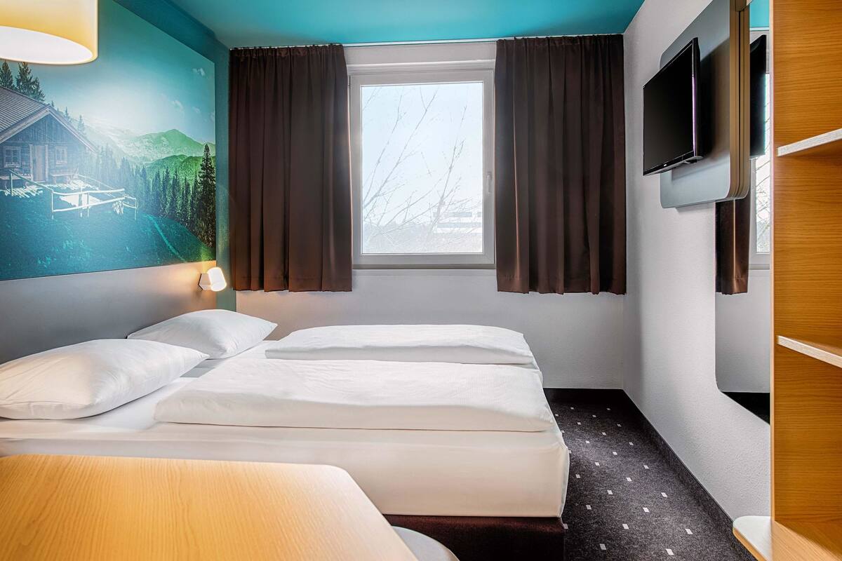 Quarto de hotel com cama, televisão presa na parede e janela com cortinas.