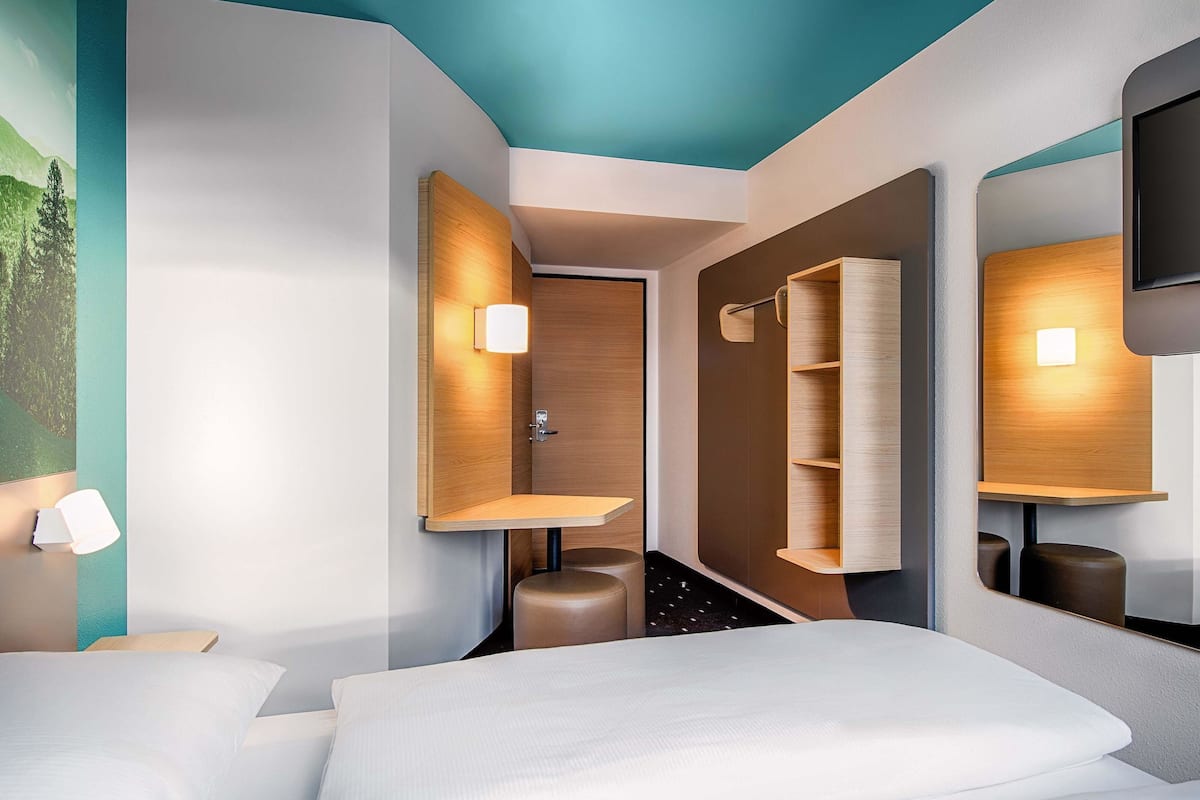 Quarto de hotel moderno com uma cama grande, um guarda-roupa de madeira, um banquinho pequeno e uma TV na parede.